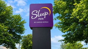 Placa Sleep Inn com fundo roxo e letras brancas, rodeada por vegetação e um céu claro.