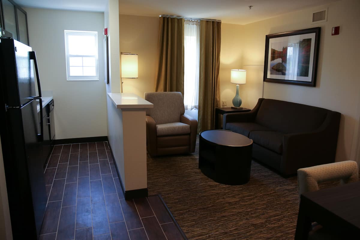 Quarto de hotel com cozinha compacta, um sofá, uma cadeira, uma mesa pequena e uma luminária.