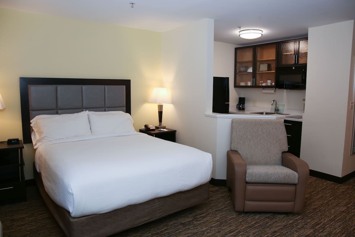 Quarto de hotel com uma cama grande, uma mesa de cabeceira com abajur, uma cadeira, uma pequena escrivaninha com telefone e uma área de cozinha compacta com micro-ondas e pia.