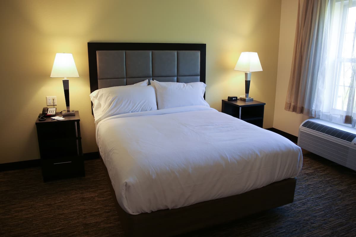 Quarto de hotel com uma cama grande, duas mesas de cabeceira com luminárias, ar condicionado na parede e uma janela com cortinas.