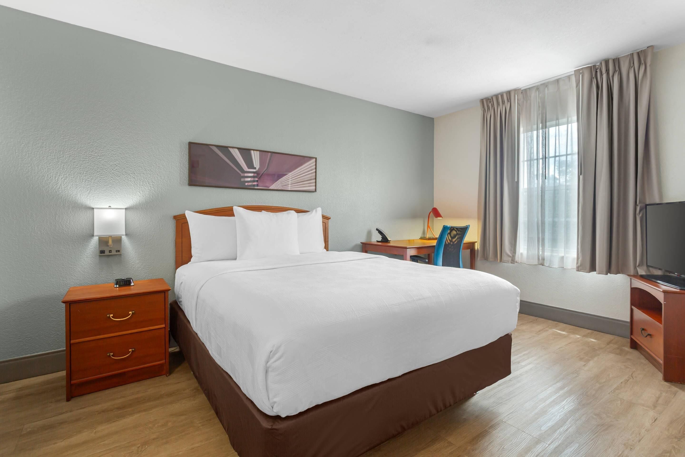 Quarto de hotel com uma cama grande, uma mesa, uma cadeira, uma TV e uma janela com cortinas.