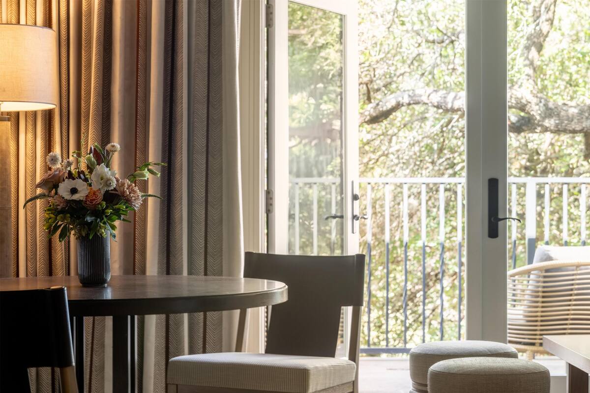 Quarto com mesa redonda, cadeiras, um vaso de flores, cortinas e vista para árvores através de uma porta de vidro.