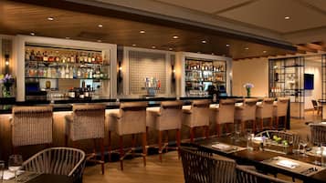 Uma área de bar com diversas opções de assento, incluindo banquetas e mesas de jantar, com variedade de garrafas e copos nas prateleiras.