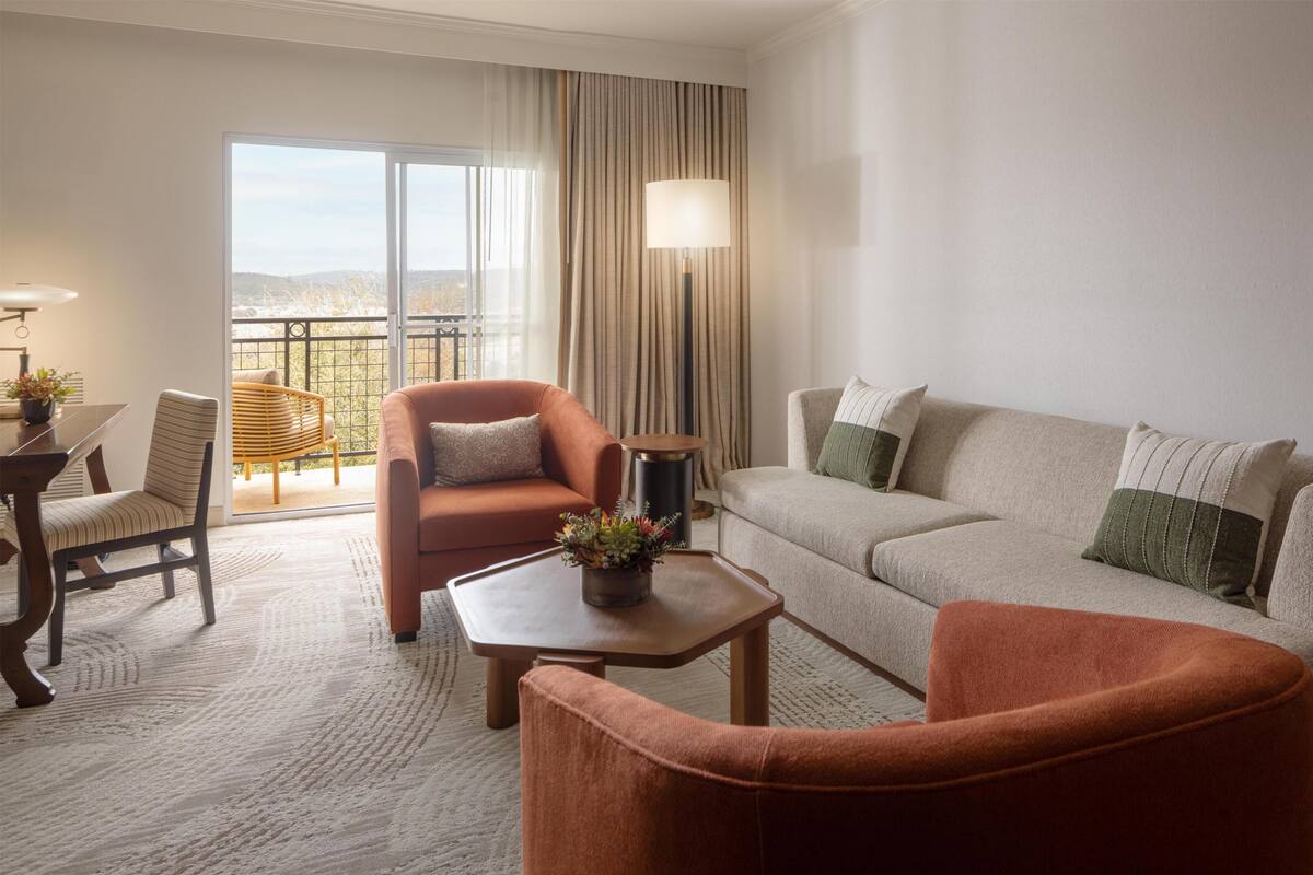 Quarto de hotel com sofá, poltrona, uma mesinha com uma planta, abajur e vista para o exterior.