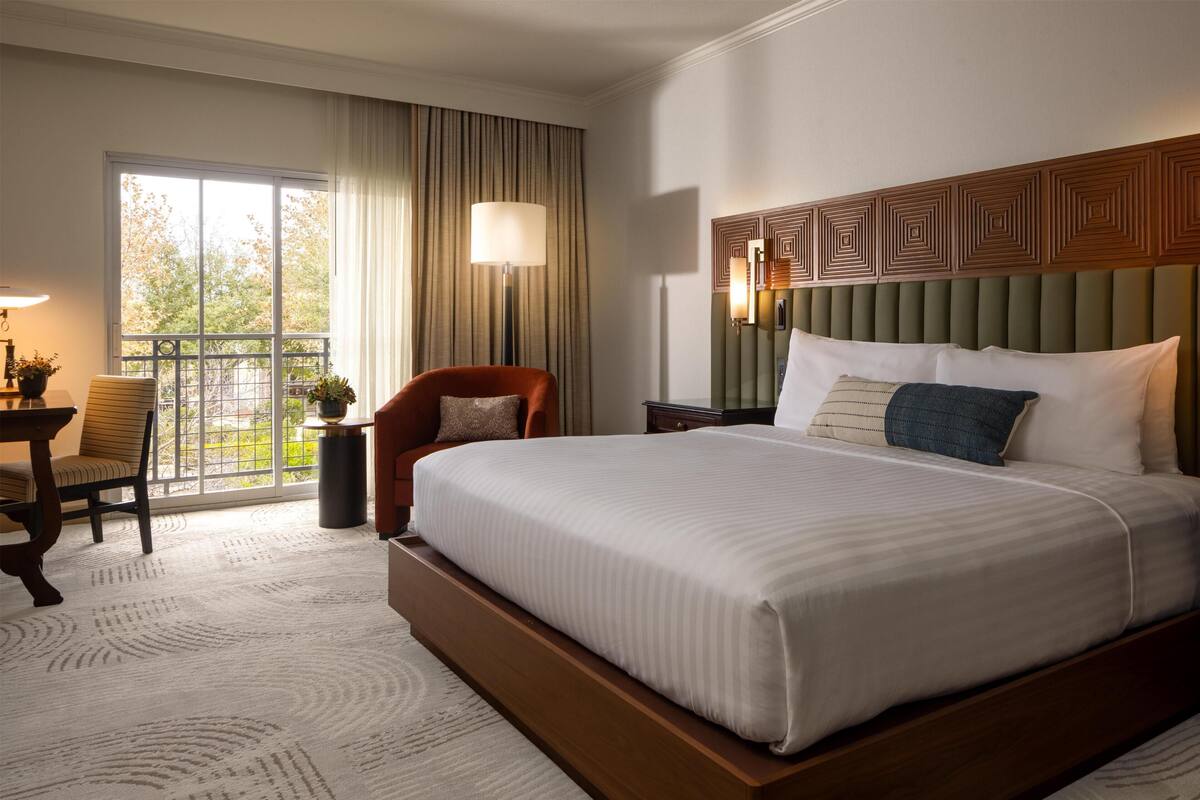 Quarto de hotel com cama grande, uma cadeira, mesa, abajur e vista para o exterior.