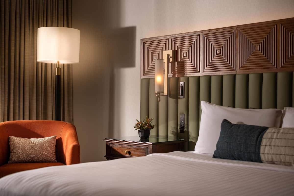 Quarto de hotel com cama, mesa de cabeceira, luminária, cadeira e cabeceira decorativa.