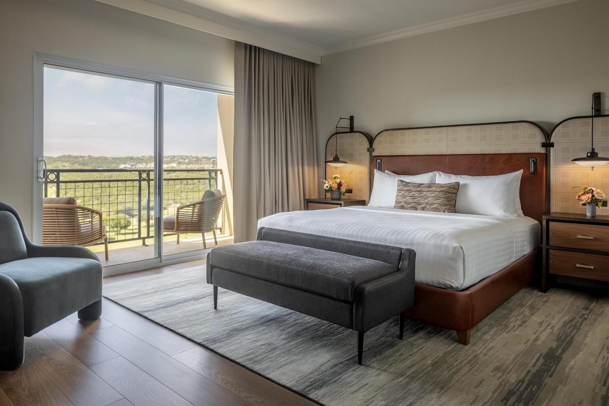 Quarto de hotel com uma cama grande, um banco, uma cadeira, uma mesa de cabeceira e vista para o exterior.