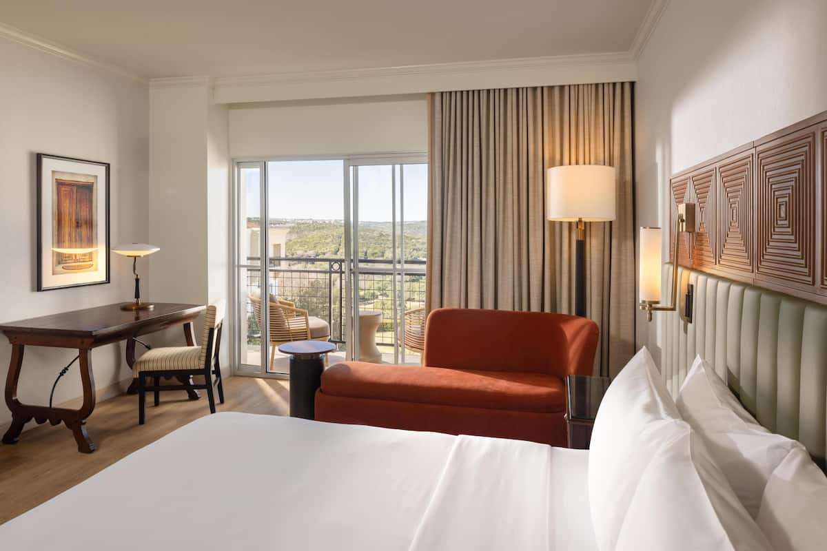 Quarto de hotel com uma cama grande, uma escrivaninha com cadeira, uma mesinha redonda pequena, uma poltrona vermelha, uma janela ampla com vista e uma luminária de parede.