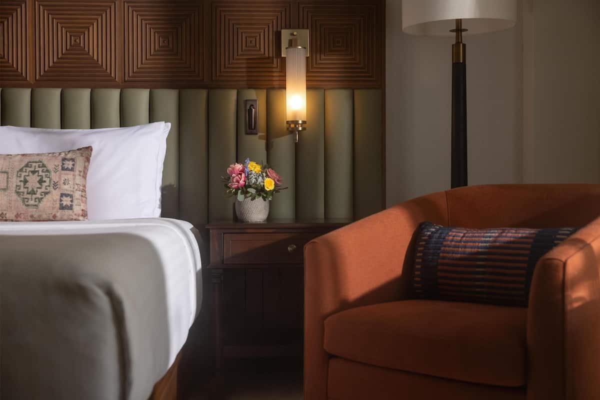 Quarto de hotel com cama, mesa de cabeceira com luminária, vaso de flores e poltrona laranja.