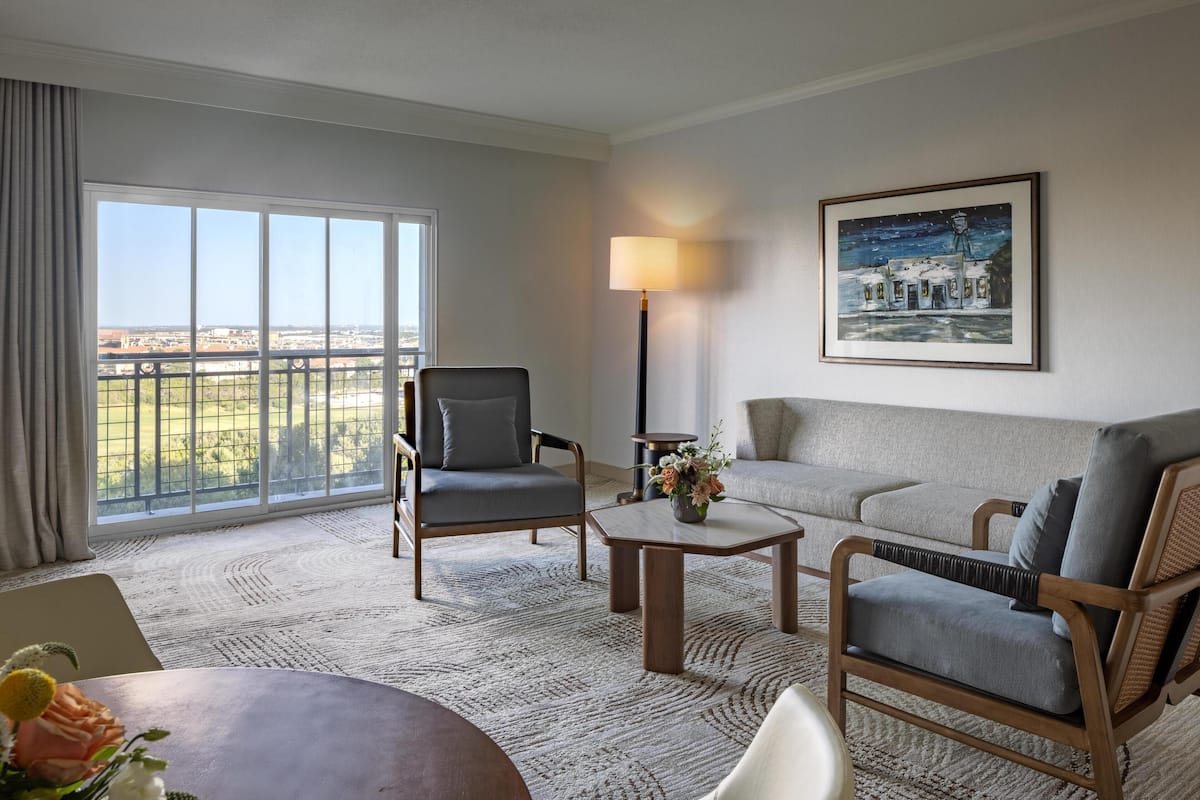 Quarto de hotel com uma grande janela, um sofá, uma cadeira, uma mesa de centro e um abajur.