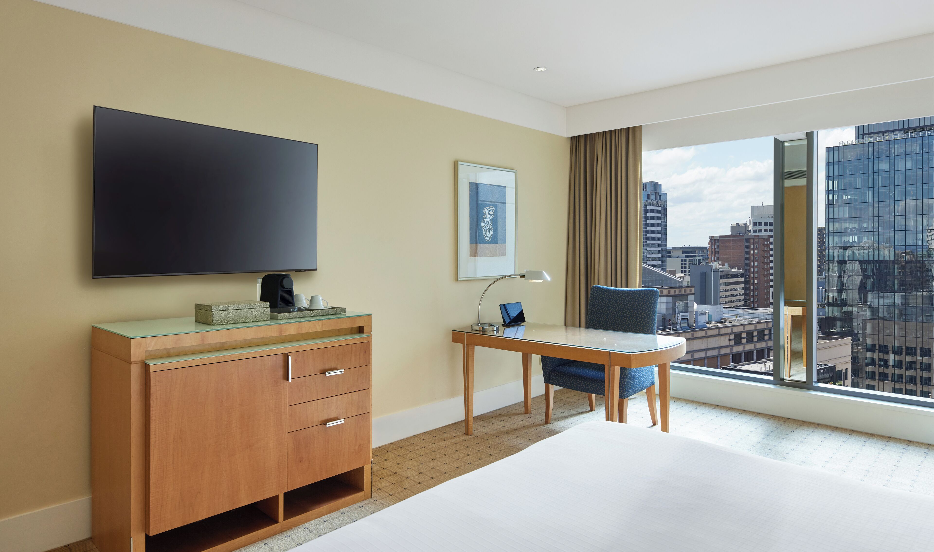 Quarto de hotel com uma televisão grande, uma escrivaninha com candeeiro, uma cadeira e vista para a cidade.