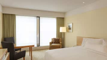 Quarto de hotel com cama, escrivaninha, duas cadeiras, abajur e janela com cortinas.