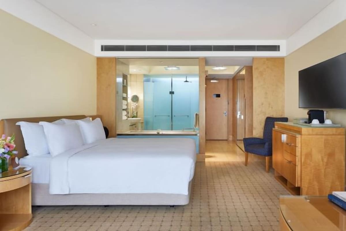 Quarto de hotel com uma cama, uma escrivaninha, uma televisão e um banheiro visível por uma porta aberta.