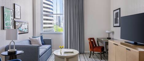 Quarto de hotel moderno com sofá cinzento, mesa de centro redonda, televisão e vista para edifícios por uma janela grande.