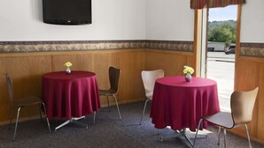 Quarto com duas mesas redondas cobertas por toalhas vermelhas, cada uma com um vaso de flores amarelas. Há quatro cadeiras, duas em cada mesa. Uma televisão de tela plana está montada na parede acima das mesas. O quarto tem um rodapé de madeira ao longo da parede inferior e uma janela com vista para um estacionamento e árvores lá fora.