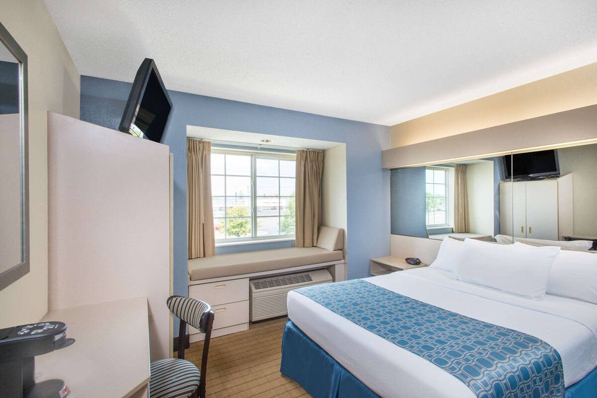 Quarto de hotel com cama, escrivaninha, cadeira, televisão e janela com cortinas.