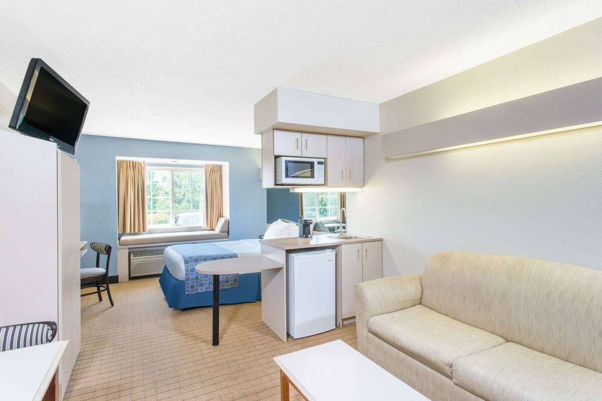 Quarto compacto com cama, cozinha compacta, sofá e televisão presa à parede.