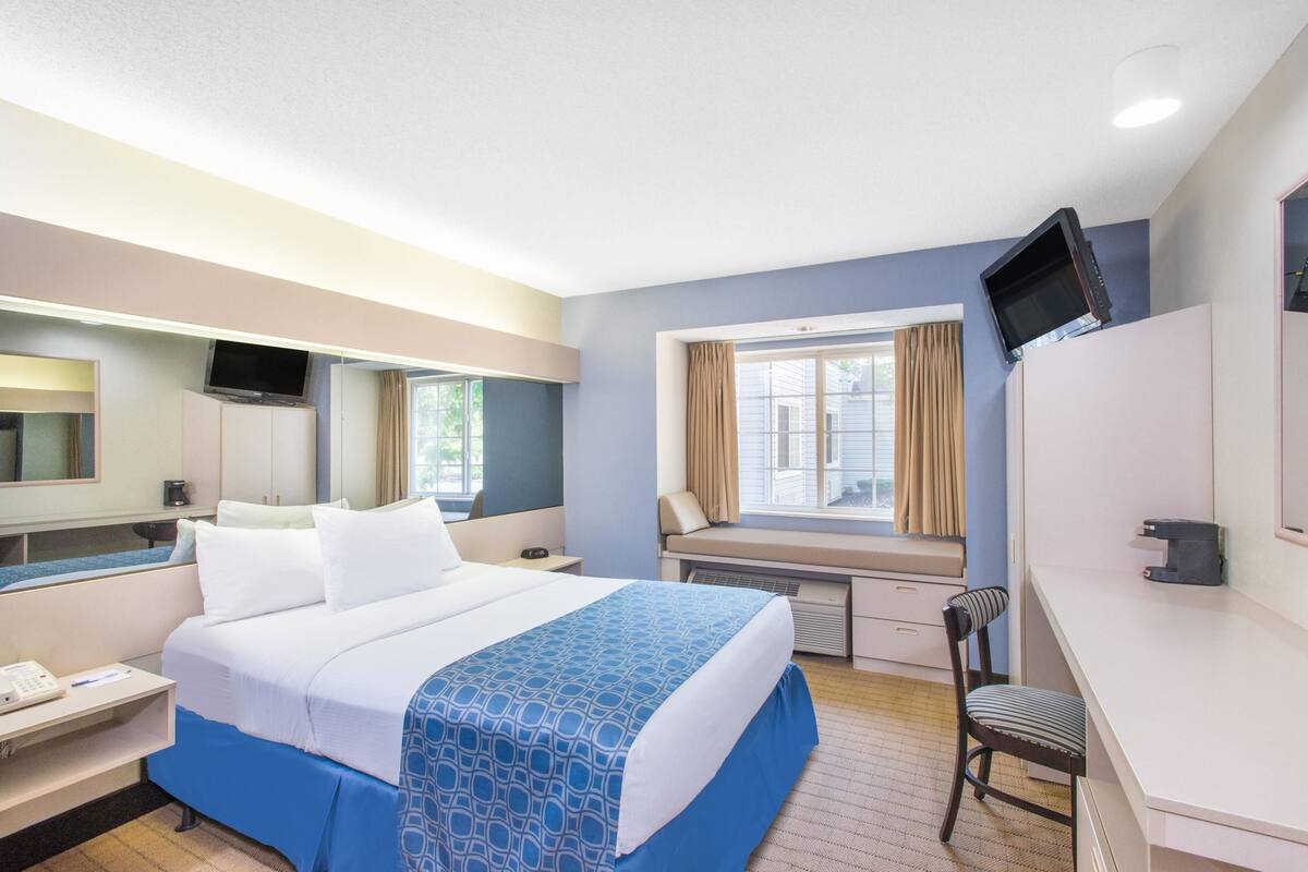 Quarto de hotel com cama, escrivaninha, cadeira, televisão e janela com cortinas.