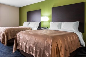 Quarto de hotel com duas camas, uma mesinha de cabeceira, uma luminária e televisão presa na parede.