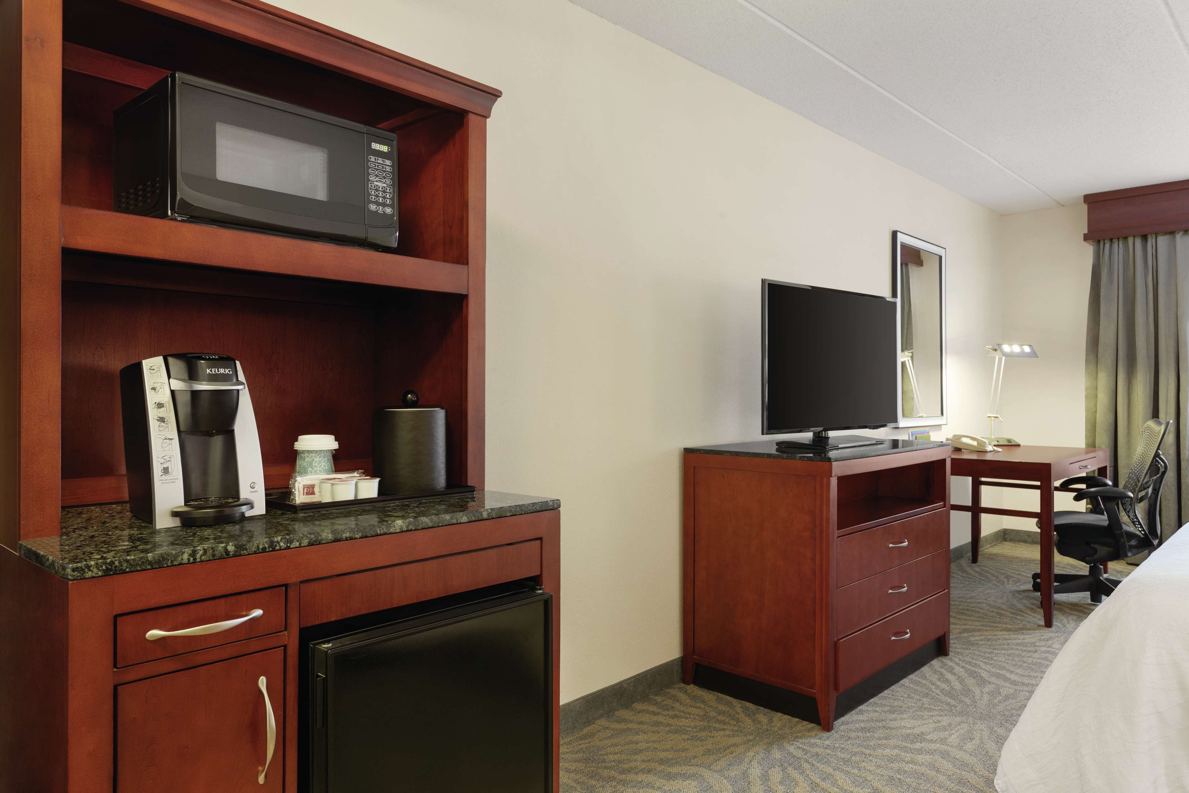 Quarto de hotel com cozinha compacta, incluindo micro-ondas, máquina de café e geladeira. Há uma mesa com computador e cadeira, e uma cama com edredom branco.
