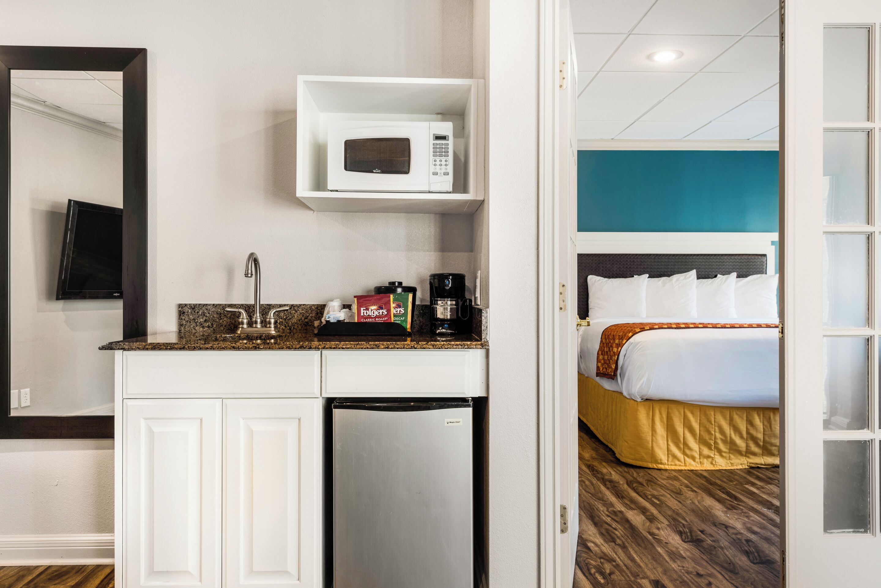 Quarto de hotel com cozinha compacta, incluindo micro-ondas, cafeteira e frigorífico. Há uma cama com roupa de cama branca e uma parede de destaque azul.