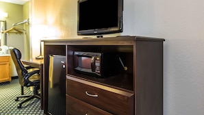 Quarto de hotel com televisão sobre um suporte de madeira, micro-ondas, escrivaninha com cadeira e cómoda.