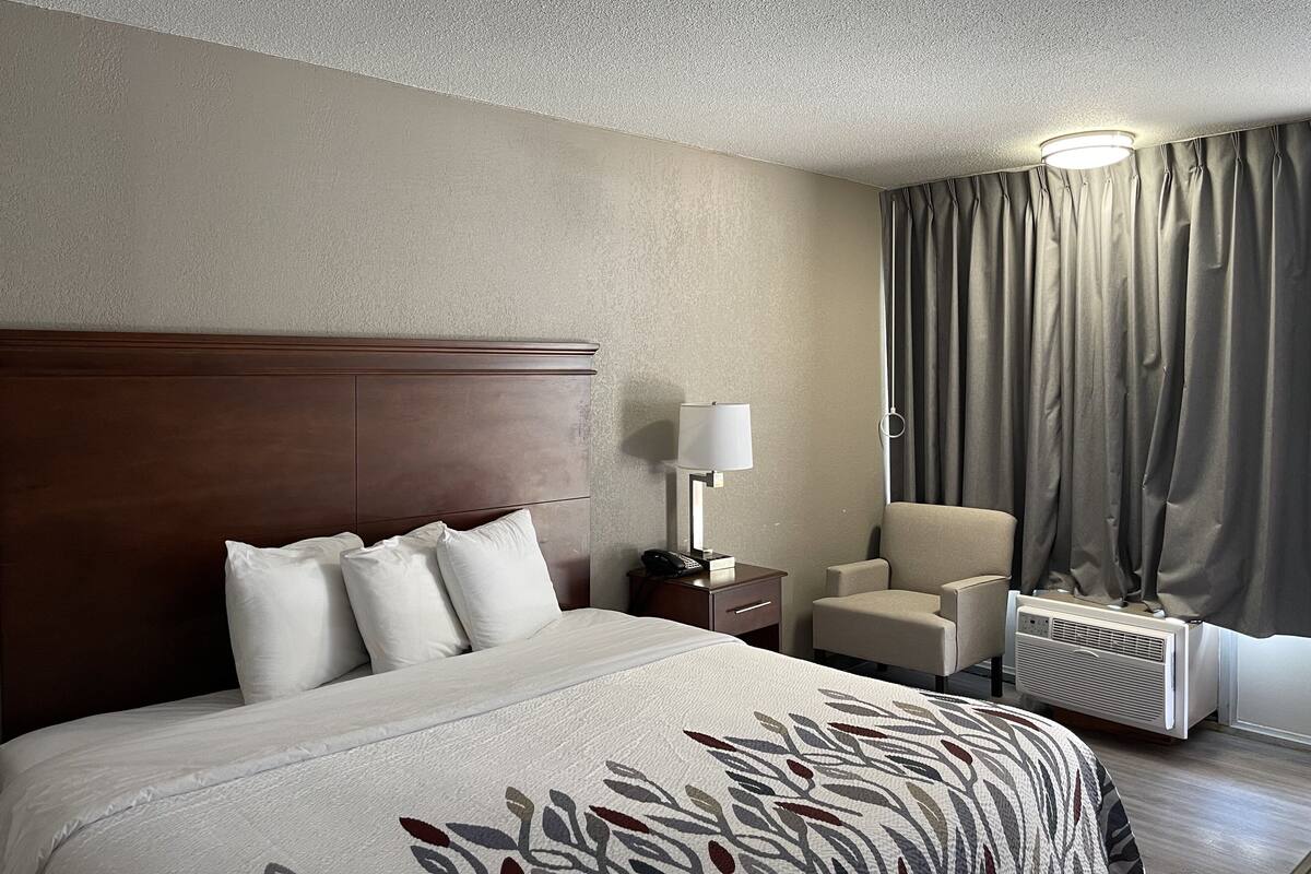 Quarto de hotel com uma cama grande, mesa de cabeceira com abajur, cadeira e janela com cortinas.