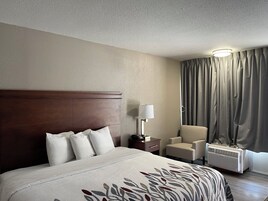 Quarto de hotel com uma cama grande, mesa de cabeceira, luminária, cadeira e janela com cortinas.