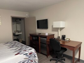 Quarto de hotel com cama, escrivaninha, cadeira, televisão, abajur e cafeteira.