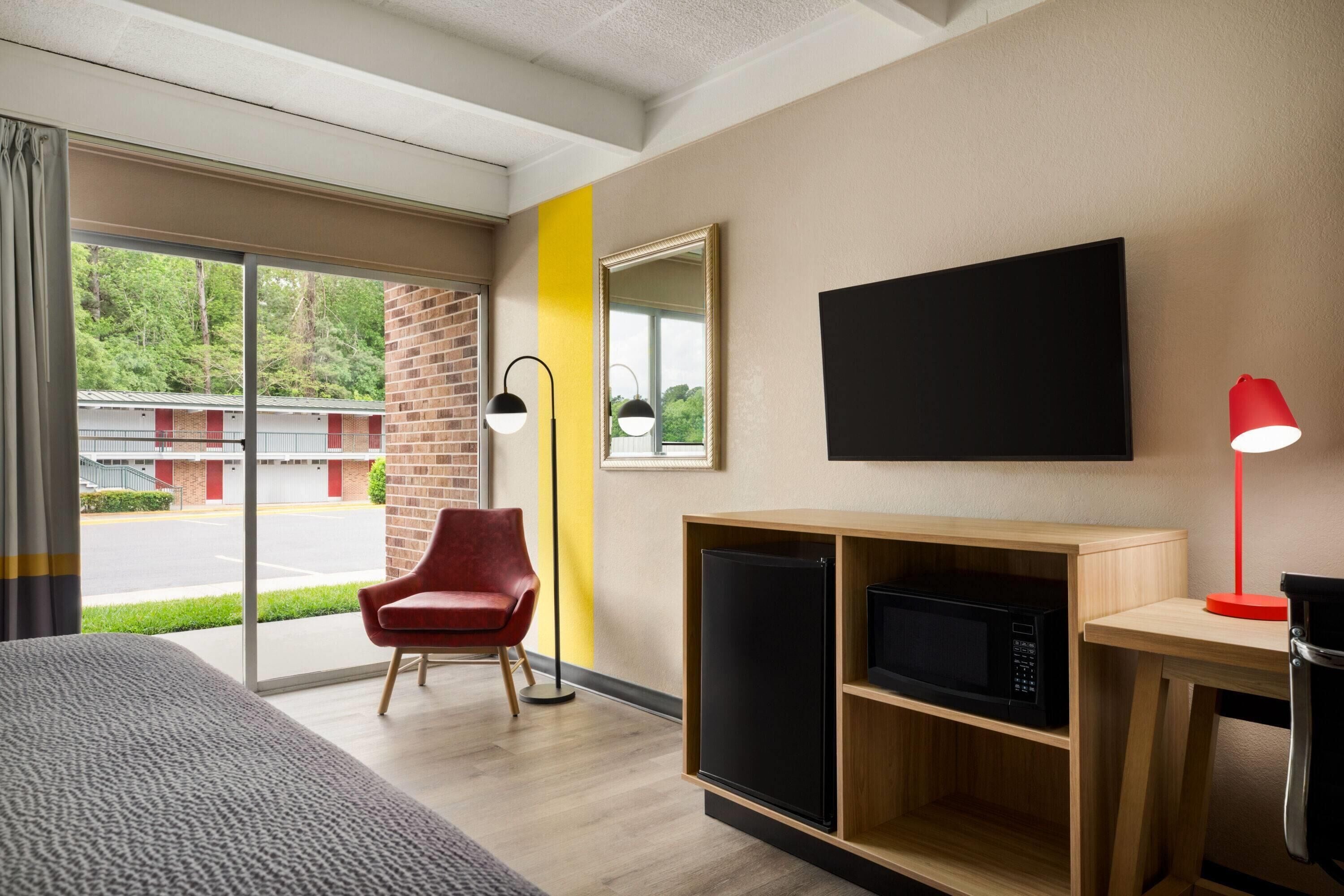 Quarto de hotel com cama, uma cadeira vermelha, televisão, micro-ondas e escrivaninha com abajur.