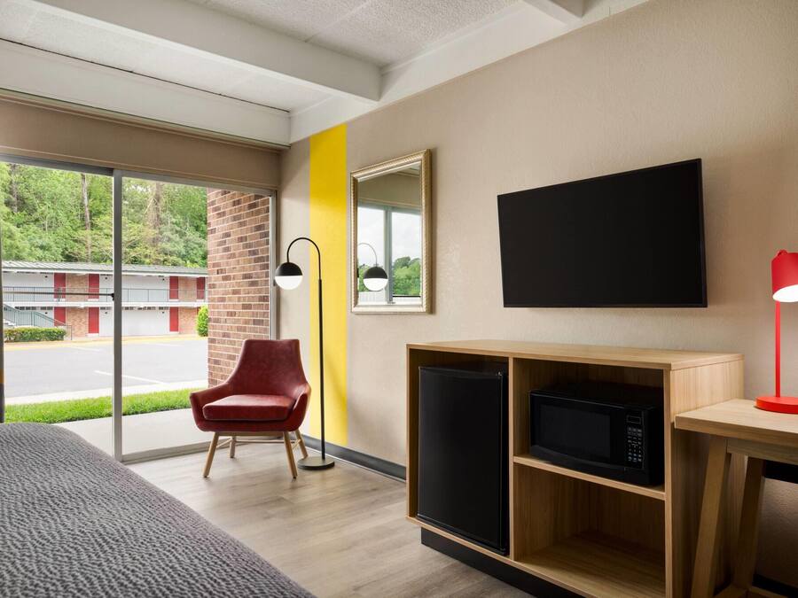 Quarto de hotel com cama, uma cadeira vermelha, televisão, micro-ondas e escrivaninha com abajur.