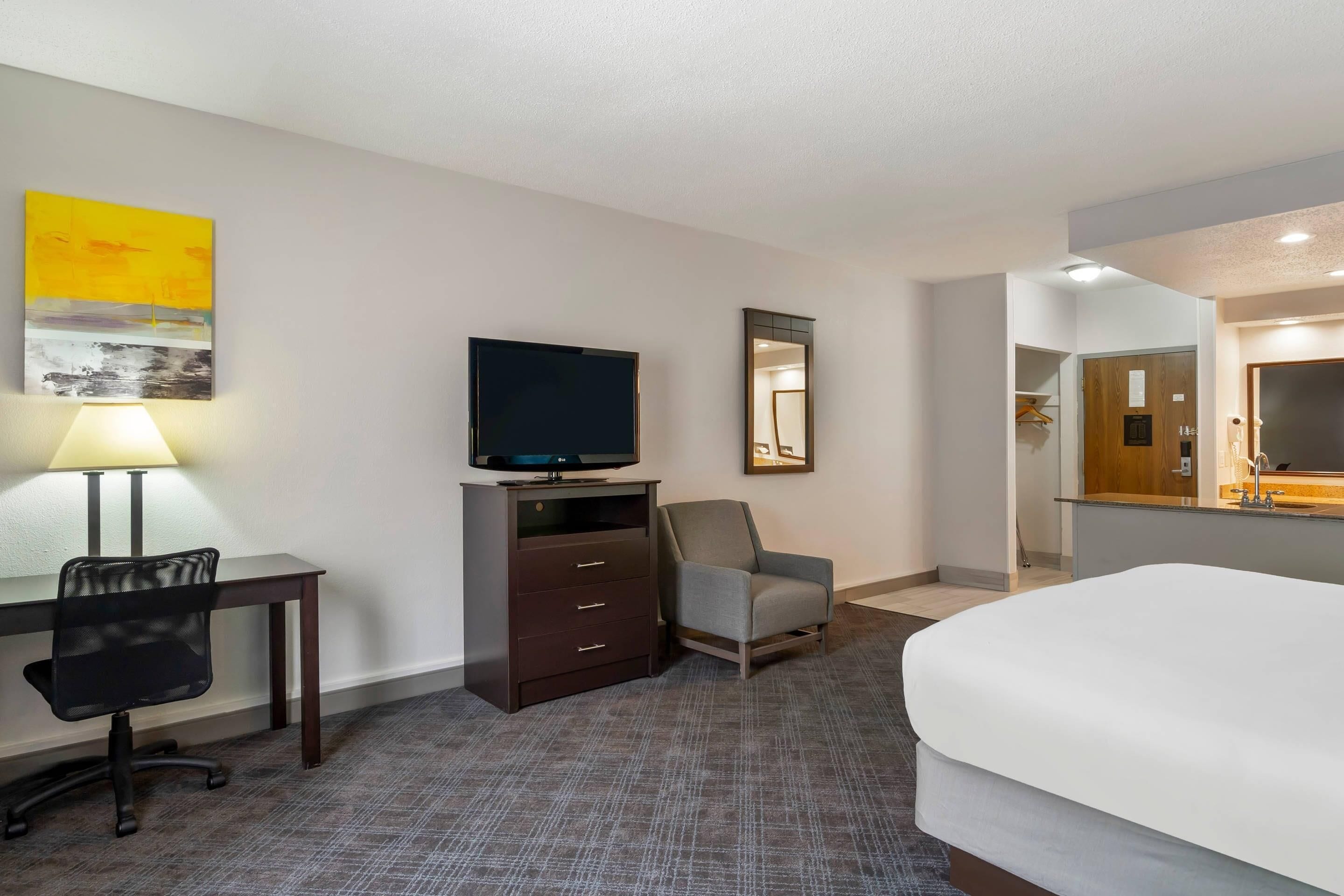 Quarto de hotel com cama, escrivaninha, cadeira, televisão, cômoda e espelho.