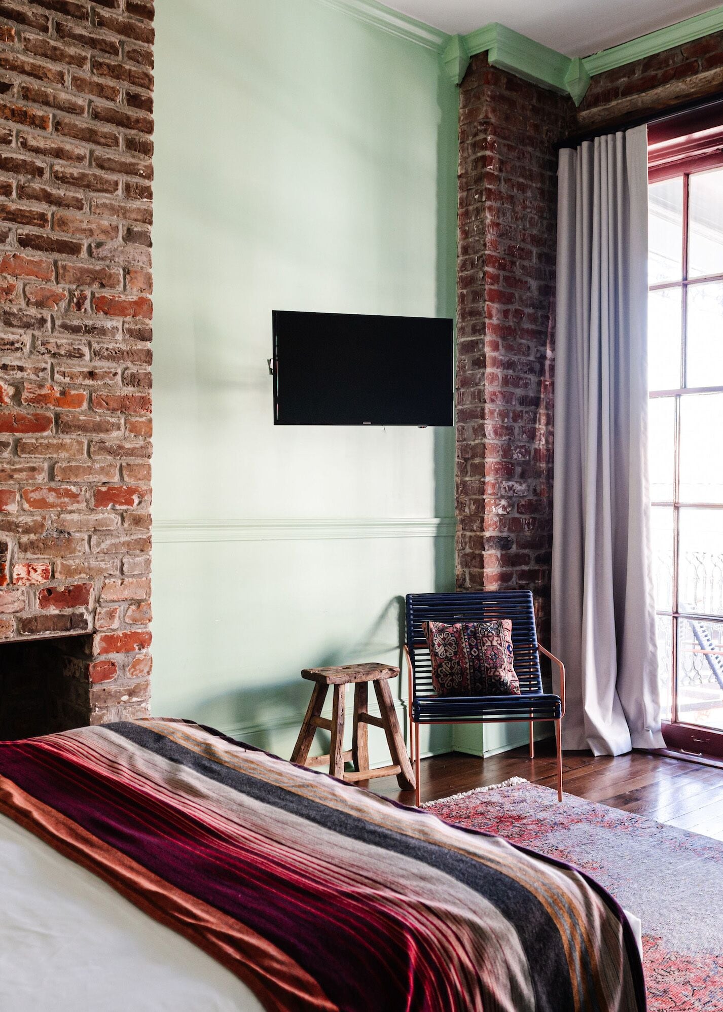 Quarto com parede de tijolos, uma TV de tela plana fixada nela, um banquinho de madeira, uma cadeira com almofada estampada e uma cama com cobertor listrado.