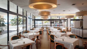 Um restaurante com várias mesas e cadeiras, luminárias pendentes grandes e amplas janelas com vista para o exterior.
