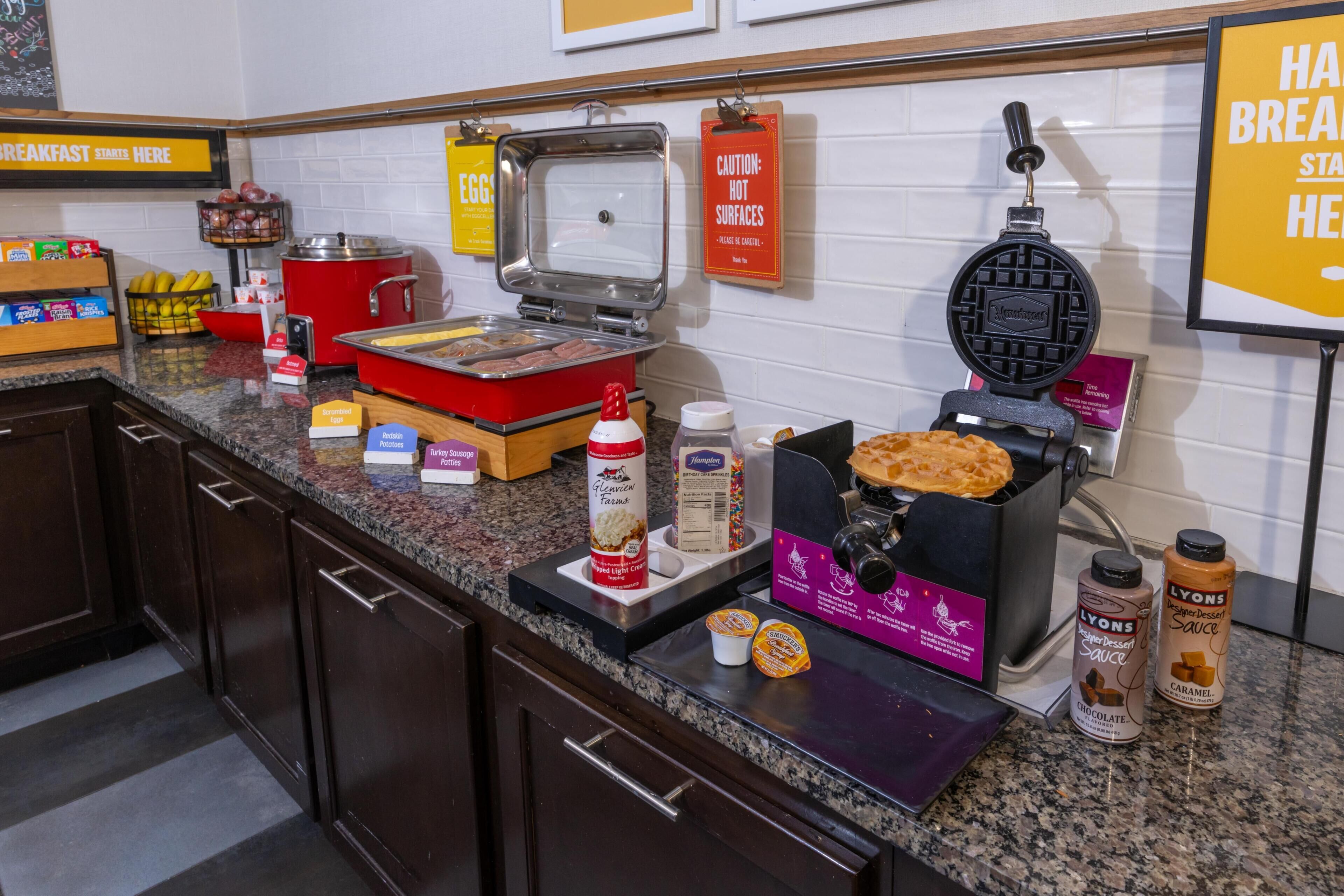 Uma mesa de café da manhã com máquina de waffles, molho picante e acompanhamentos.