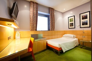 Quarto de hotel com uma cama, uma escrivaninha, uma cadeira, uma TV e duas molduras na parede.