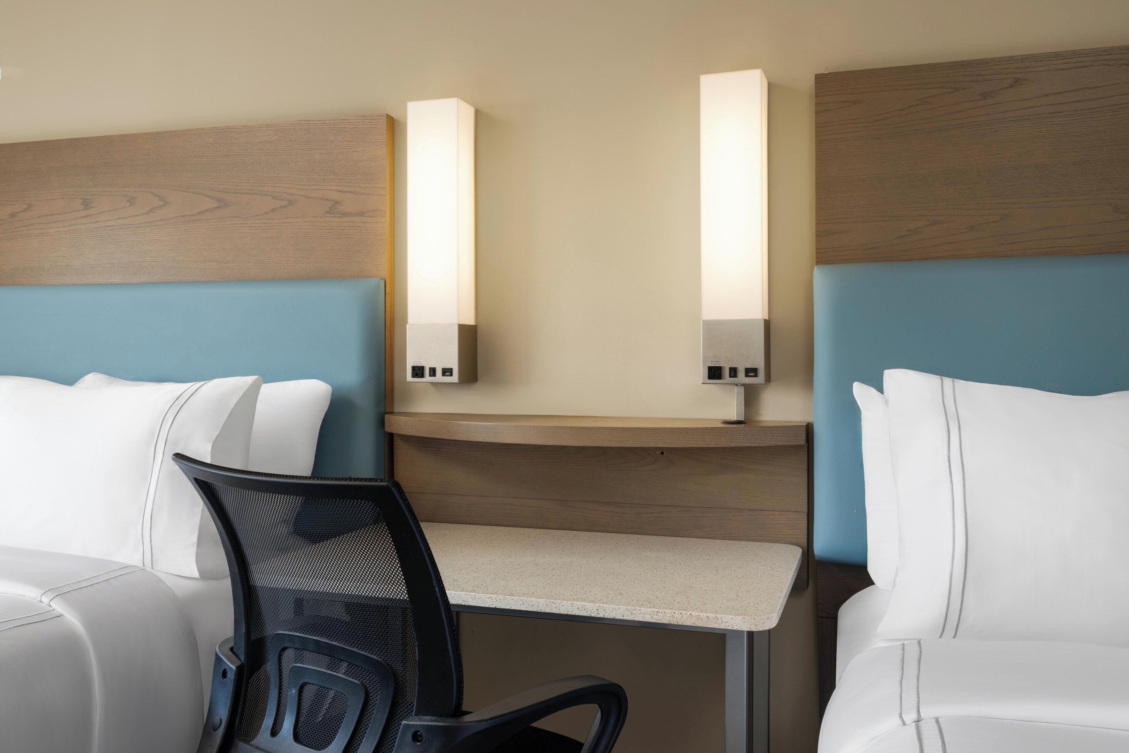 Quarto de hotel com duas camas, uma escrivaninha e duas luminárias fixadas na parede.