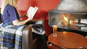 Uma pessoa lendo um livro em um quarto aconchegante, com lareira e uma mesa de madeira redonda com um copo de cerveja.
