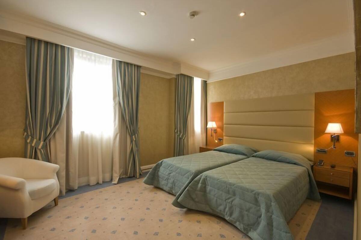 Quarto de hotel com duas camas, uma cadeira e uma mesinha de cabeceira com abajur.