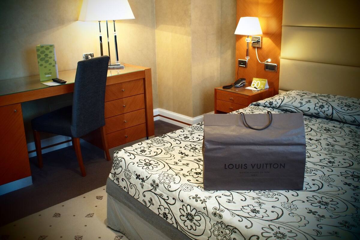 Quarto de hotel com escrivaninha, cadeira, mesa de cabeceira, abajur e uma cama com colcha estampada. Uma sacola de compras Louis Vuitton está colocada sobre a cama.