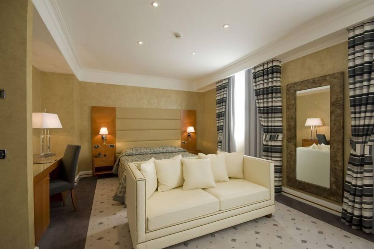 Quarto de hotel com cama, sofá, escrivaninha com luminária, espelho e cortinas.