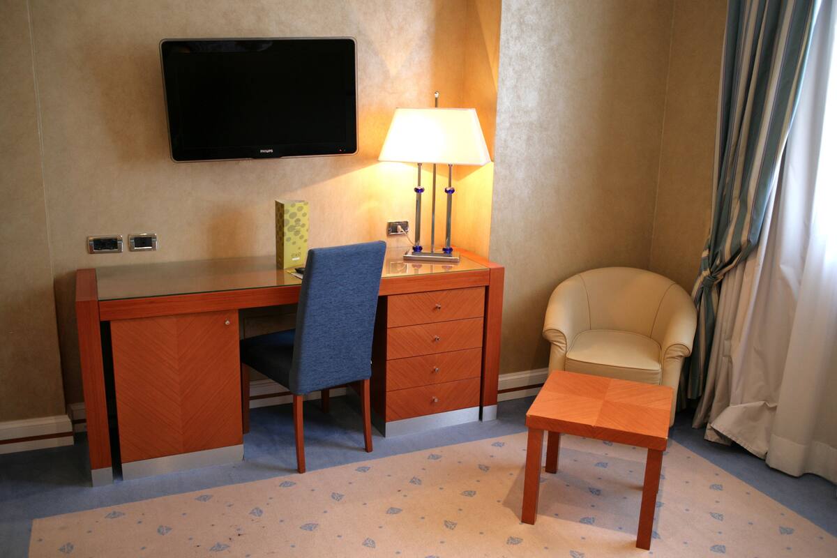 Quarto de hotel com escrivaninha, cadeira, televisão, luminária e uma pequena mesa.