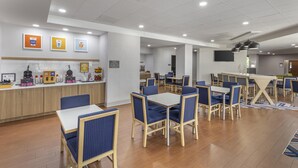 Uma sala de jantar moderna com mesas de madeira e cadeiras estofadas em azul, uma bancada de cozinha com diversos eletrodomésticos e quadros nas paredes.