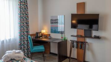 Quarto de hotel com cama, escrivaninha com cadeira, televisão, mesinha e janela com cortinas.