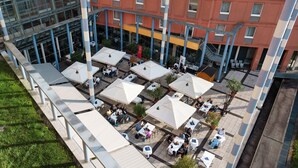 Área externa de café com mesas e guarda-sóis, cercada por um edifício moderno com varandas.