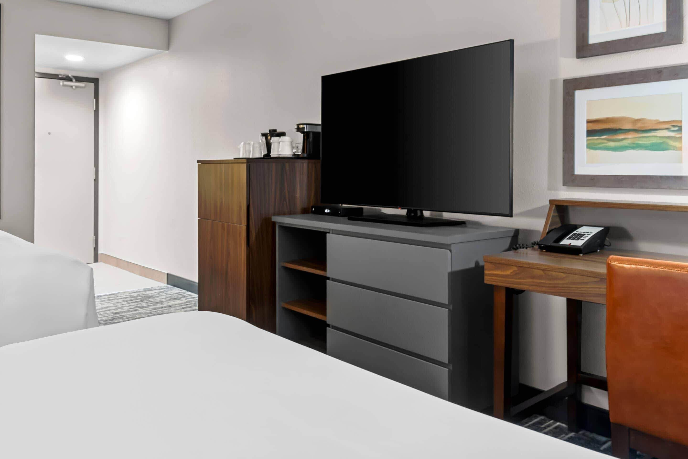 Quarto de hotel moderno com televisão de tela plana presa a uma escrivaninha de madeira, uma cama, um guarda-roupa e um pequeno espaço de estar.