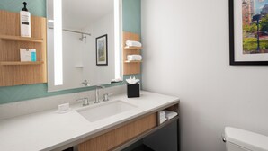 Uma casa de banho moderna com um espelho grande, uma sanita branca e uma casa de banho. Existem prateleiras de madeira com artigos de higiene pessoal e uma moldura com um quadro na parede.