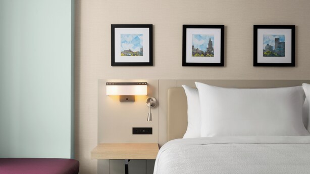 Quarto de hotel com cama, mesa de cabeceira com luminária e três quadros na parede.