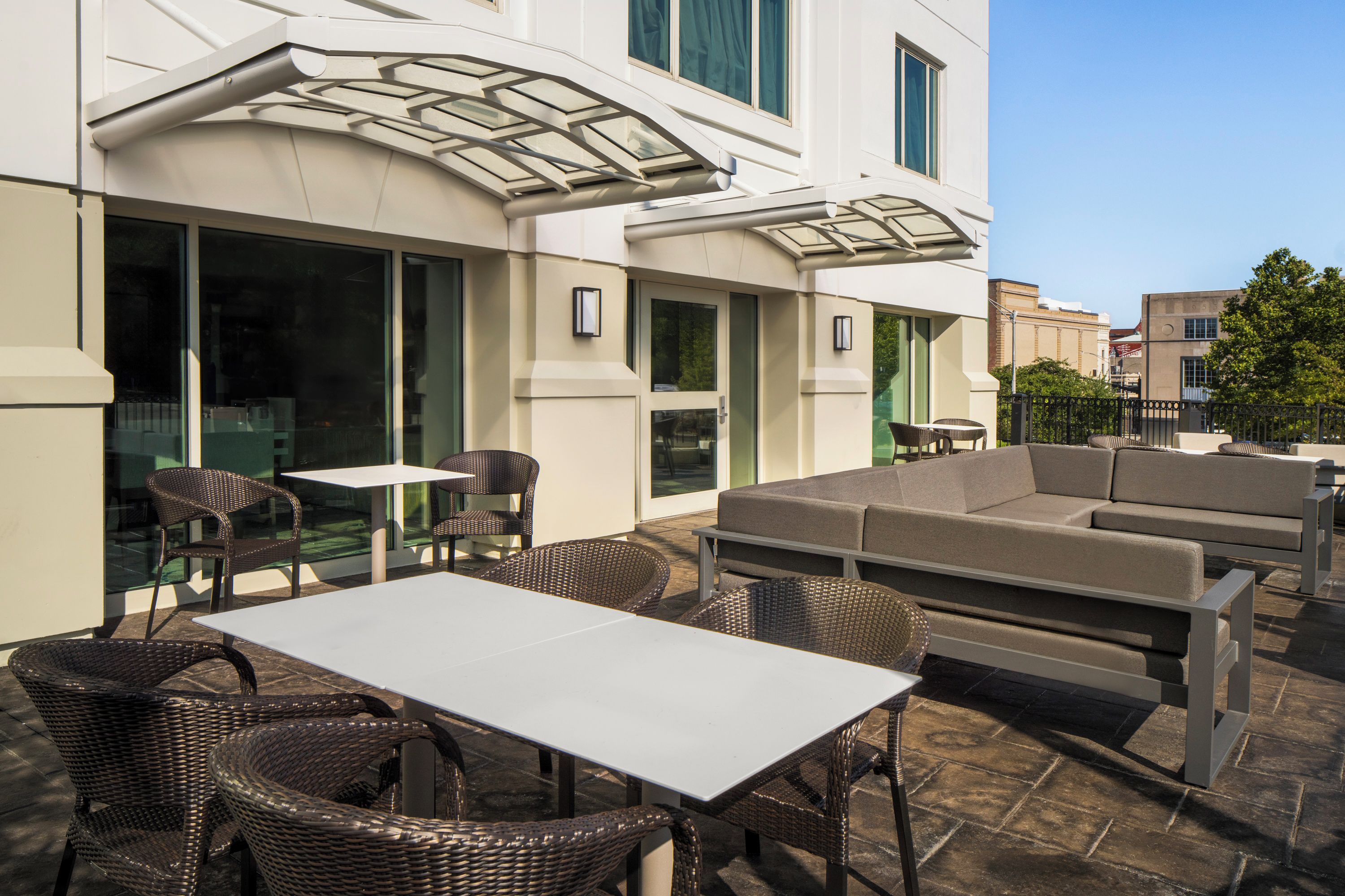 Área de estar externa com móveis de vime e uma mesa branca, adjacente a um edifício com janelas amplas e um terraço coberto.