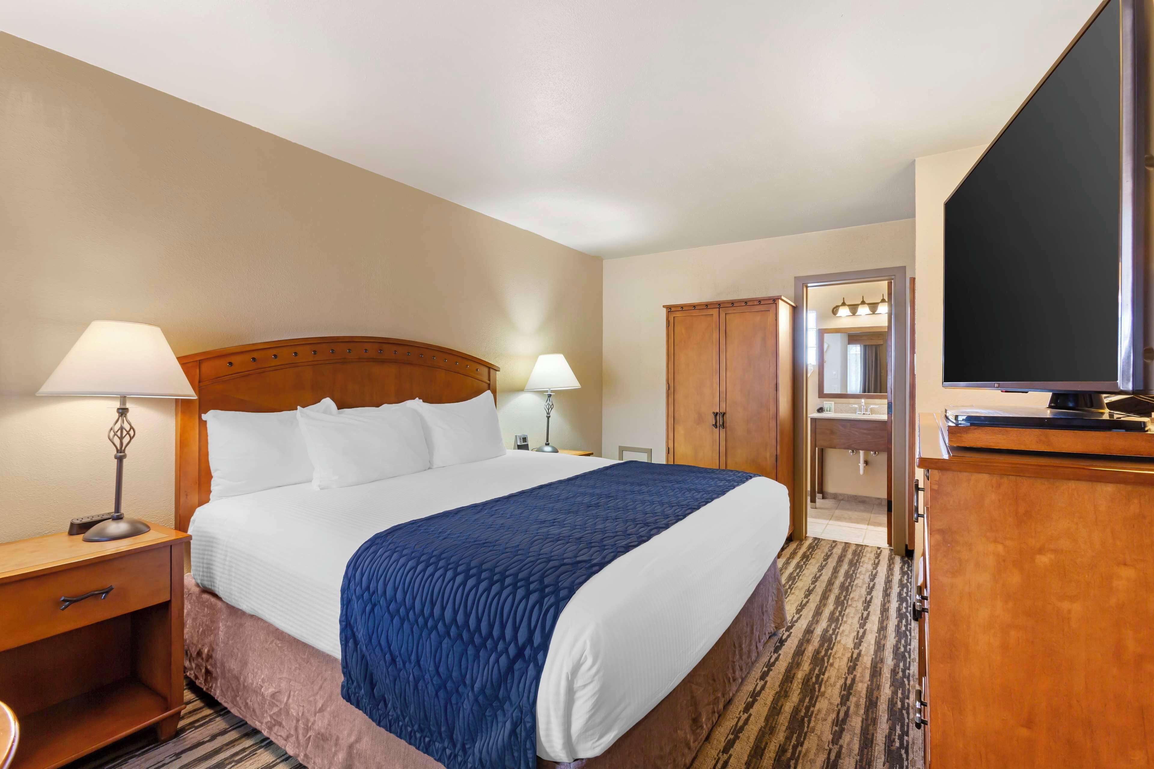 Quarto de hotel com uma cama, luminárias de cabeceira, televisão, guarda-roupa e um banheiro visível através de uma porta aberta.